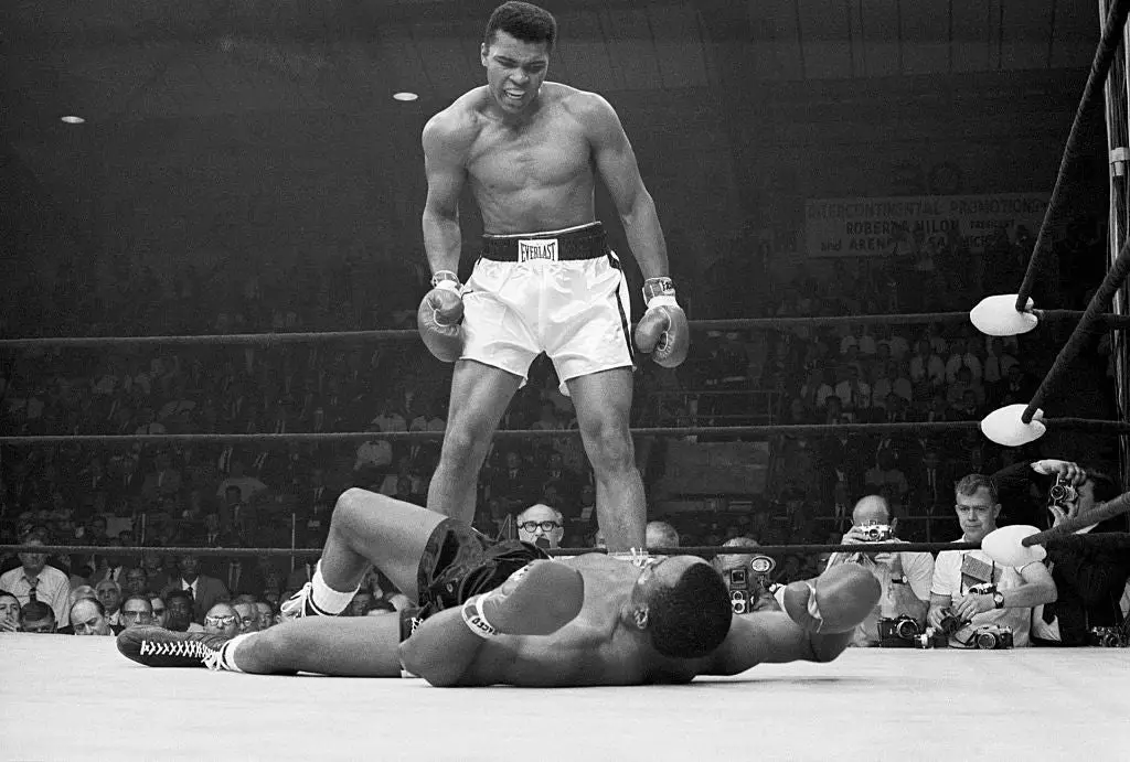 Þungavigtarmeistarinn Muhammad Ali stendur yfir Sonny Liston og grínir hann til að standa upp í titilbardaga þeirra. Ali sló Liston út á einni mínútu í fyrstu lotu í bardaga þeirra í Central Maine ungmennamiðstöðinni í Lewiston, Maine.