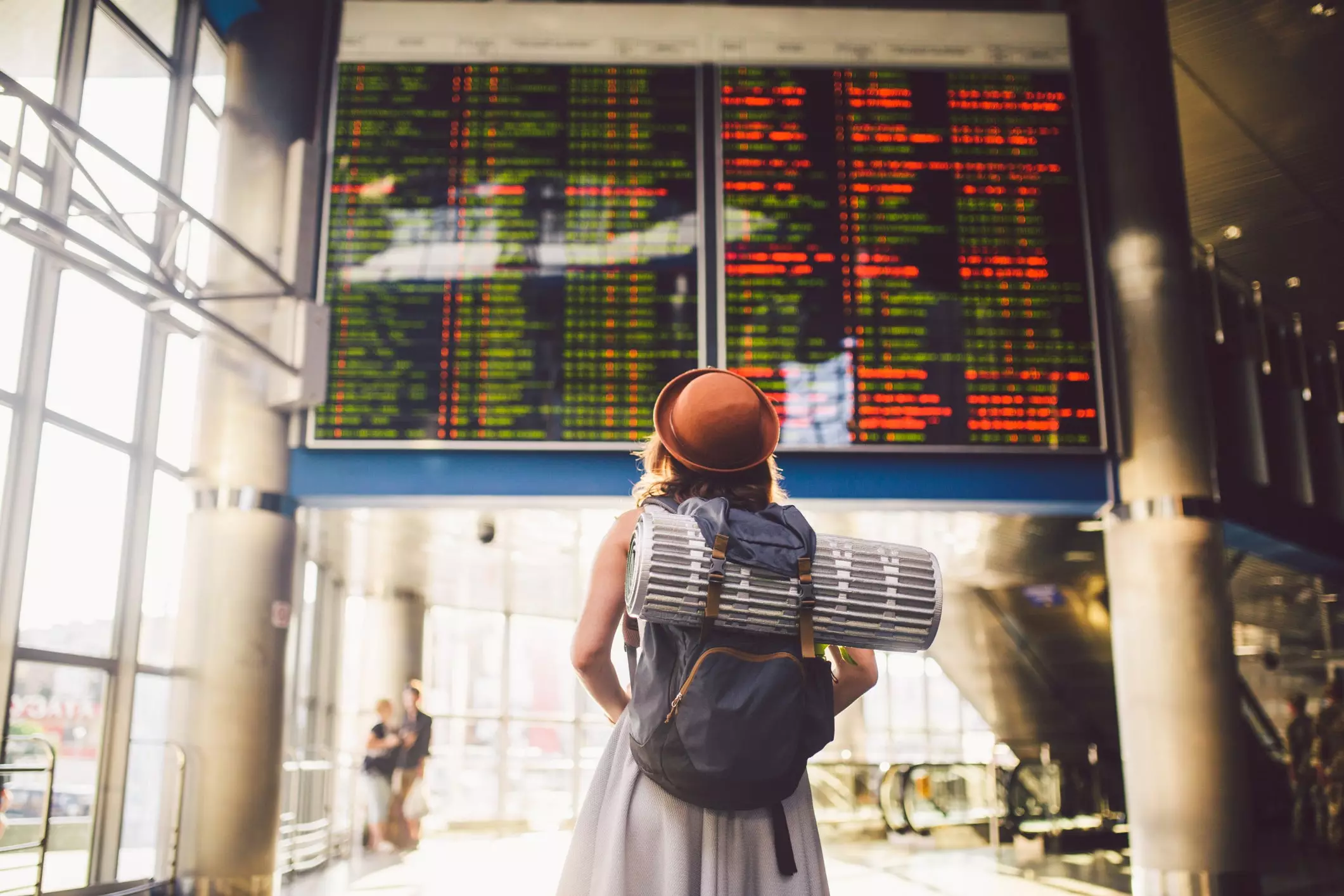 Thema Reisen öffentlicher Verkehr. junge frau, die mit rücken in kleid und hut hinter rucksack und campingausrüstung zum schlafen steht, isoliermatte sieht zeitplan auf anzeigetafel flughafenbahnhof aus